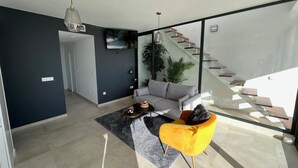 Uma sala de estar moderna com um sofá, uma poltrona amarela, uma mesa de centro e uma escadaria visível através de uma porta de vidro.