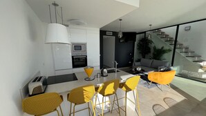 Cozinha moderna com ilha central, sala de jantar com cadeiras amarelas e sala de estar com sofá e escada.