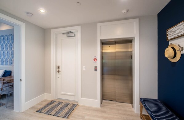 Um elevador moderno com acabamento metálico, uma porta branca com puxador e um pequeno botão vermelho de emergência.