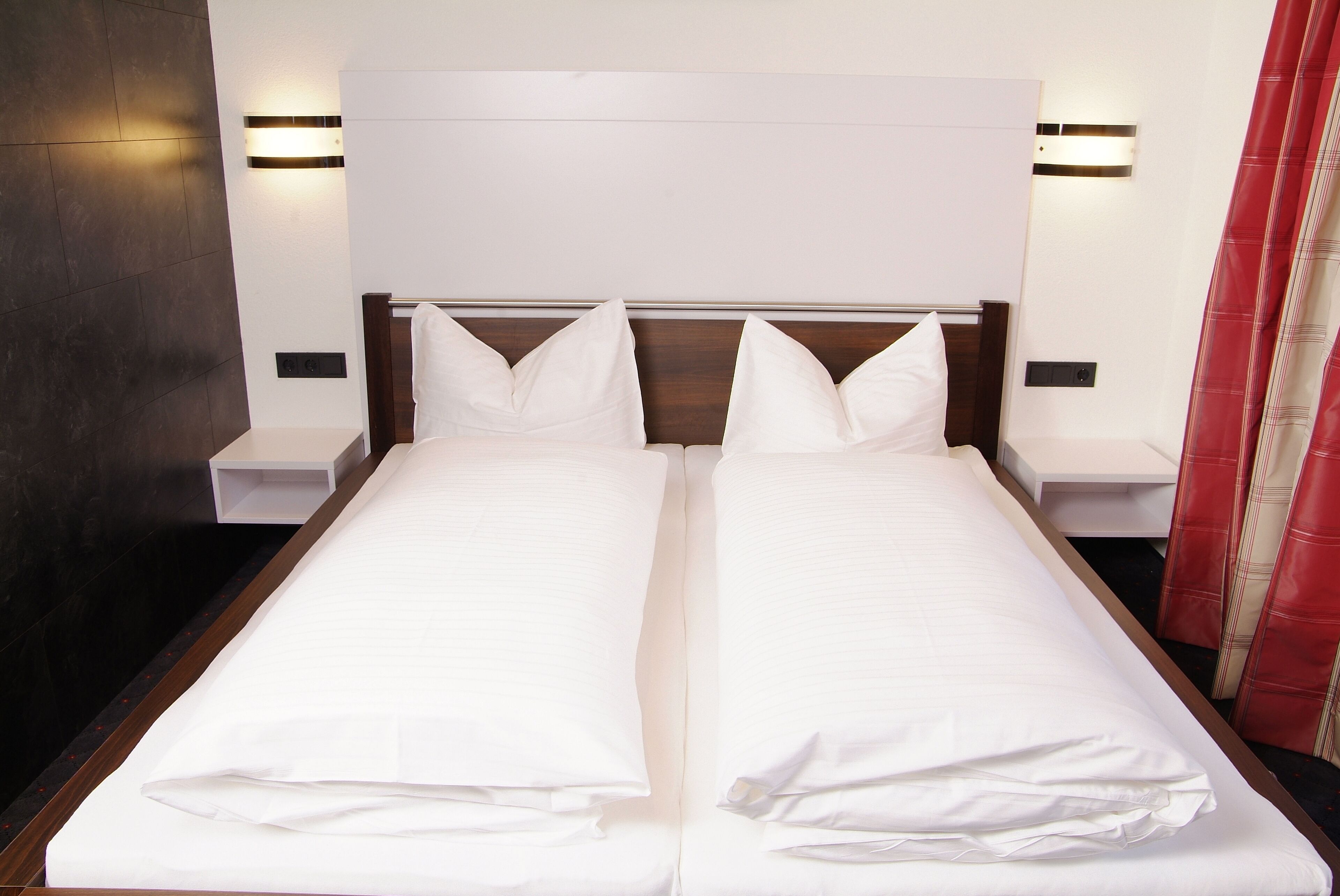 Uma cama de casal com lençóis brancos, duas mesas de cabeceira e luminárias fixadas na parede.