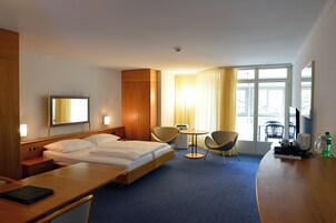 Quarto de hotel com cama, escrivaninha, cadeira, mesinha, televisão e janela com cortinas.