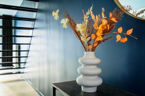 Um vaso branco com plantas secas sobre uma mesa escura em um quarto com parede azul e uma janela com persianas.