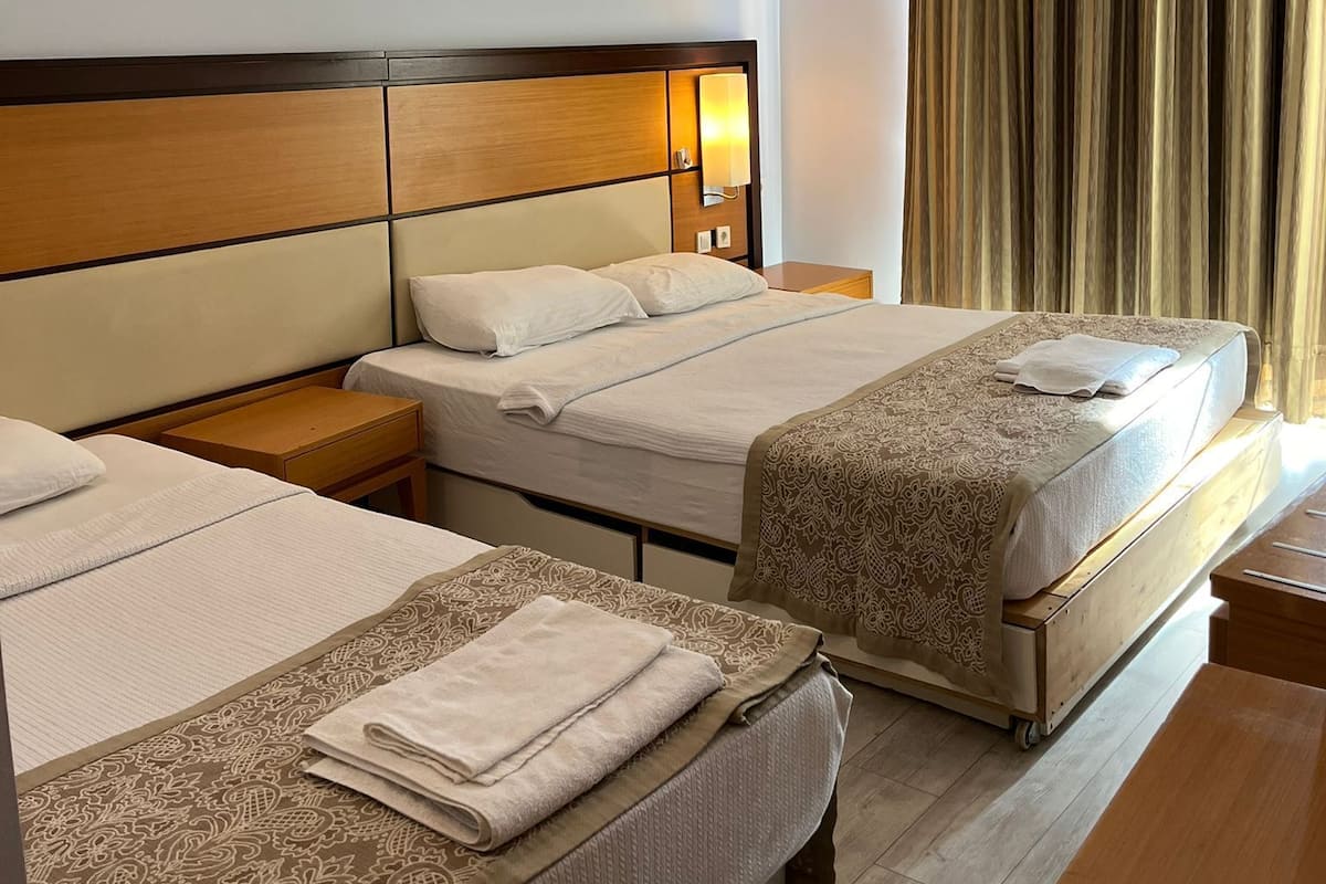 Quarto de hotel com duas camas, cabeceira de madeira, mesa de cabeceira, luminária e janela com cortinas.