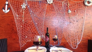 Mesa posta com dois pratos, cada um com uma toalha dobrada e um garfo, uma taça de vinho e uma garrafa de vinho. A mesa está posta em frente a uma parede vermelha com textura, com uma rede decorativa suspensa.