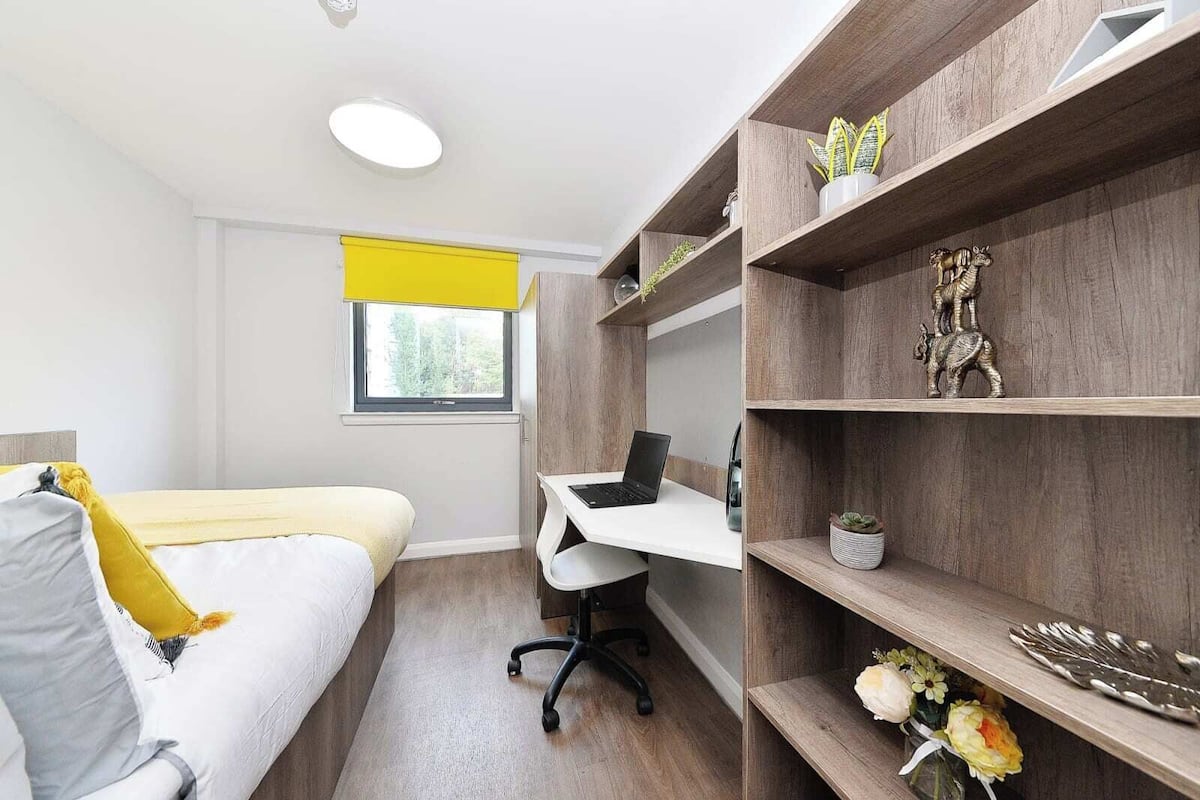 Quarto moderno com cama, escrivaninha e computador. Há uma janela com persiana amarela, uma prateleira com objetos decorativos e uma pequena planta em vaso.