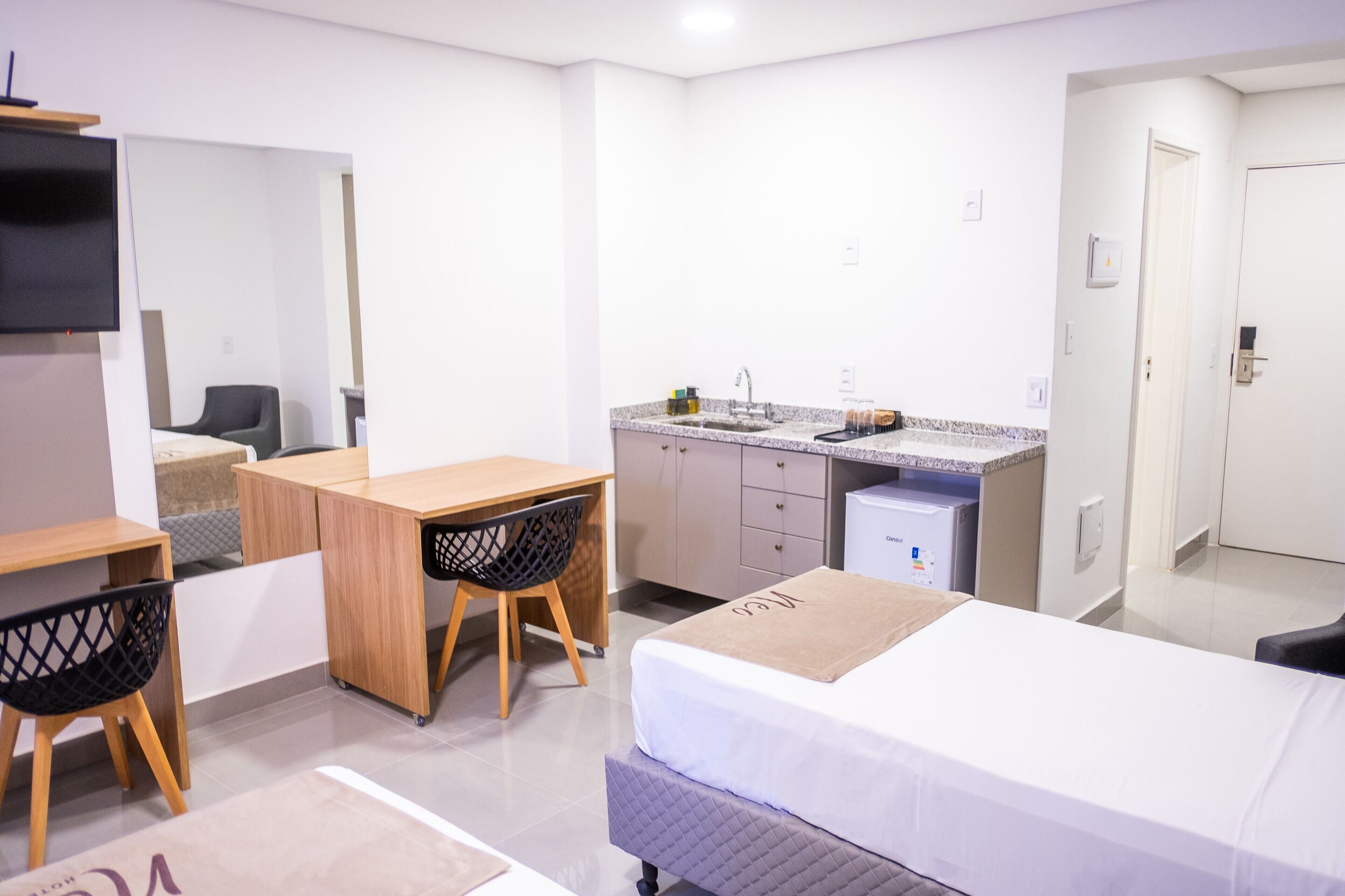 Quarto de hotel com cama, escrivaninha, cadeira, televisão, cozinha compacta e banheiro.