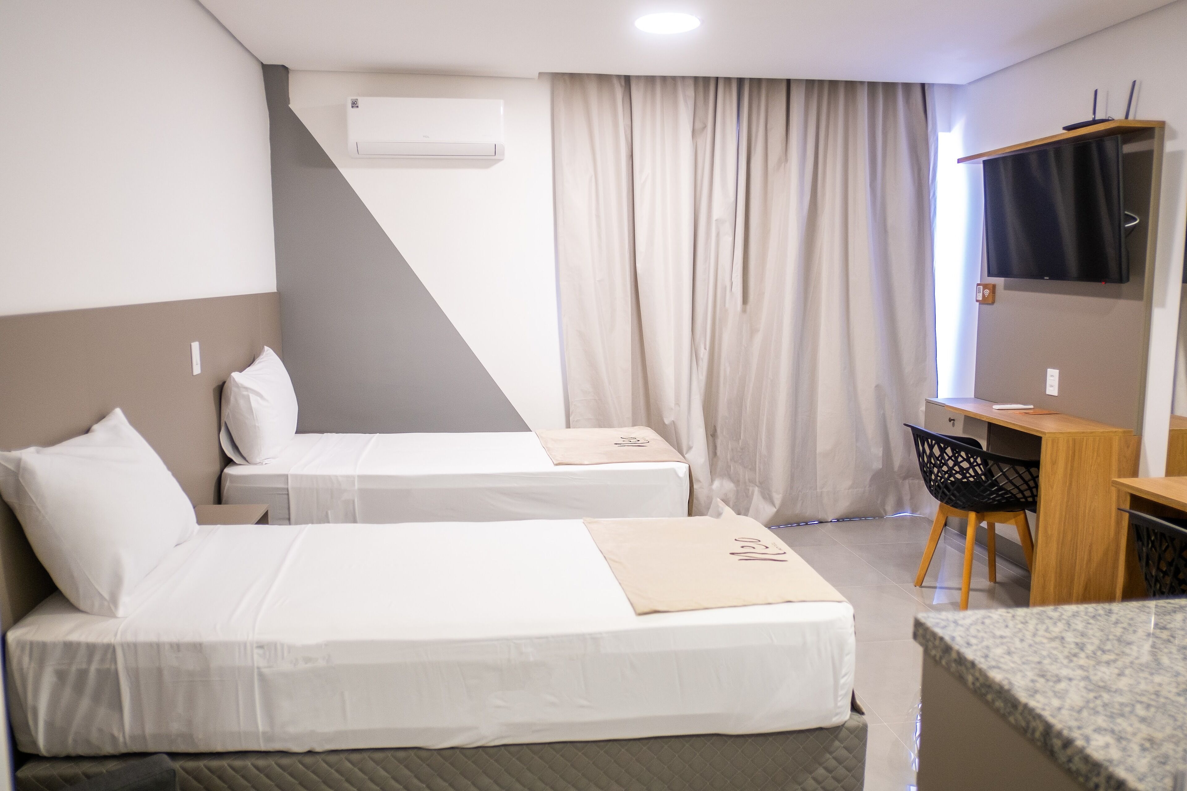 Quarto de hotel com duas camas de solteiro, uma escrivaninha, uma cadeira e uma TV fixada na parede.