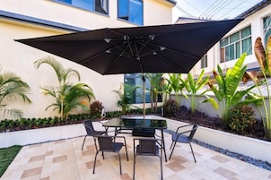 Um pátio externo com mesa, cadeiras, um guarda-sol grande e um vaso de plantas.