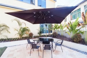 Pátio externo com mesa e cadeiras, cercado por plantas e um edifício.