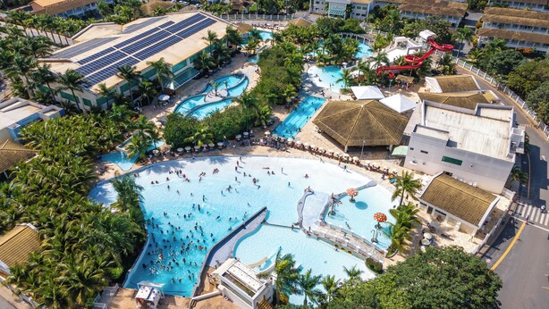 Vista aérea de um resort com várias piscinas, palmeiras e um tobogã vermelho.