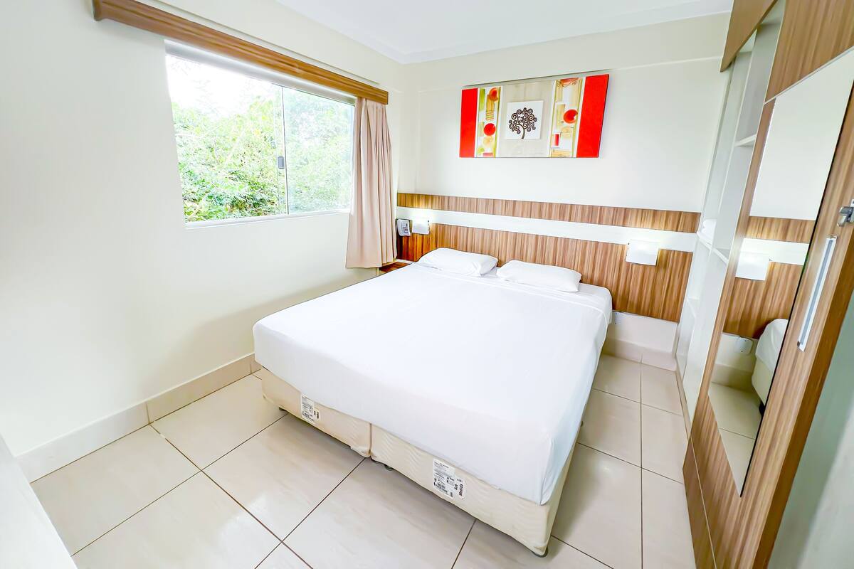 Quarto de hotel com cama grande, janela com cortinas, cabeceira de madeira e um ornamento na parede.