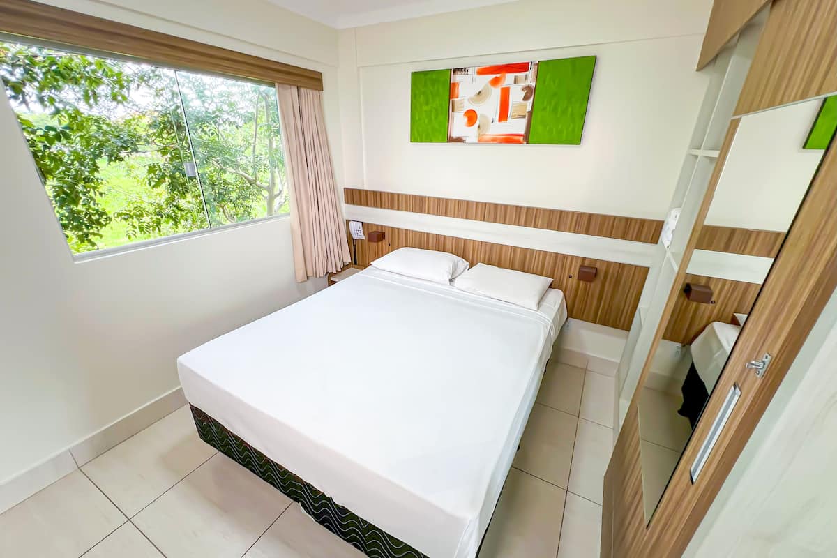 Quarto de hotel com cama, janela com cortinas, obra de arte na parede e guarda-roupa de madeira.