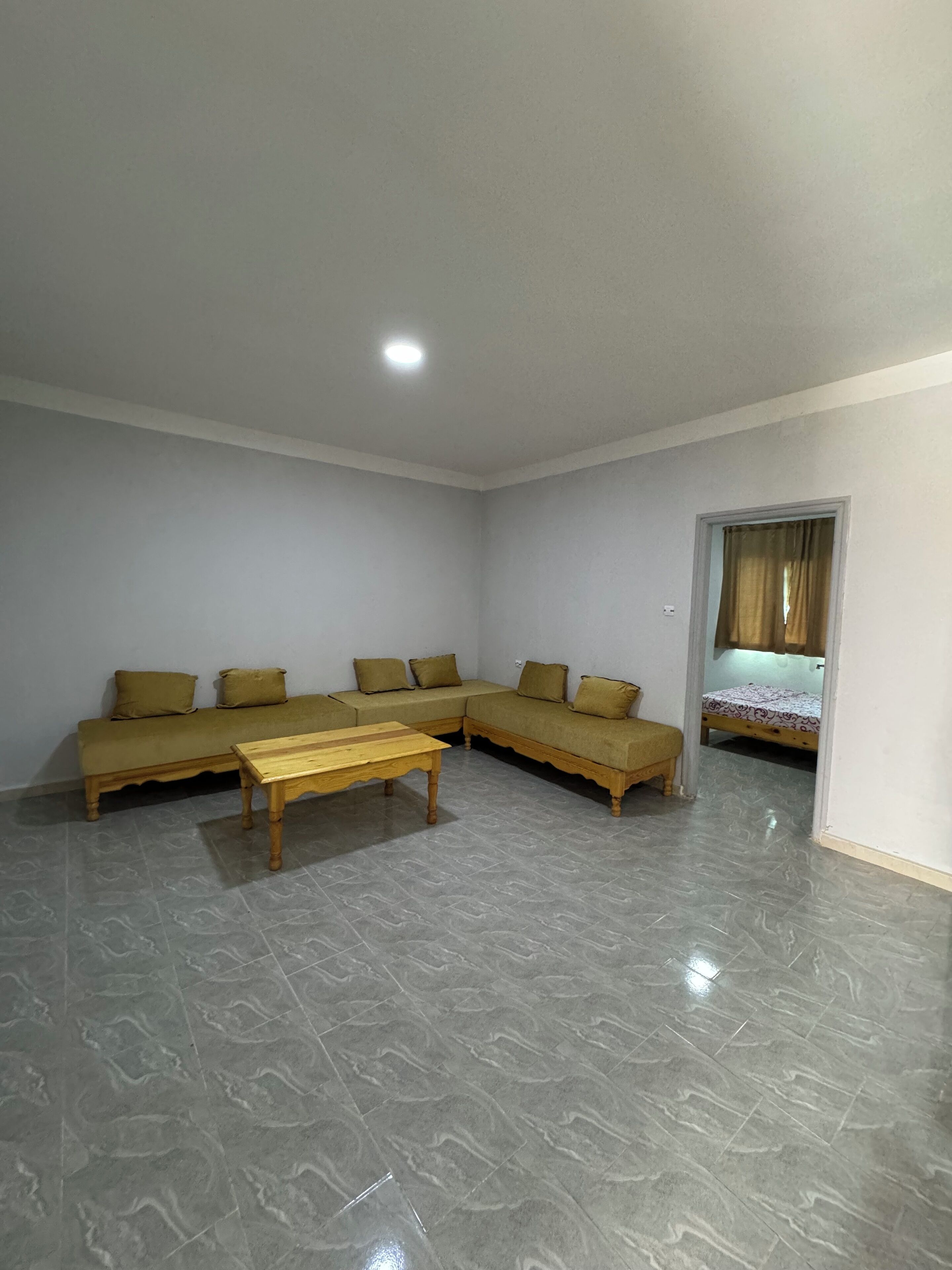 Uma sala de estar com sofá seccional, mesa de centro de madeira e uma cama no quarto adjacente.