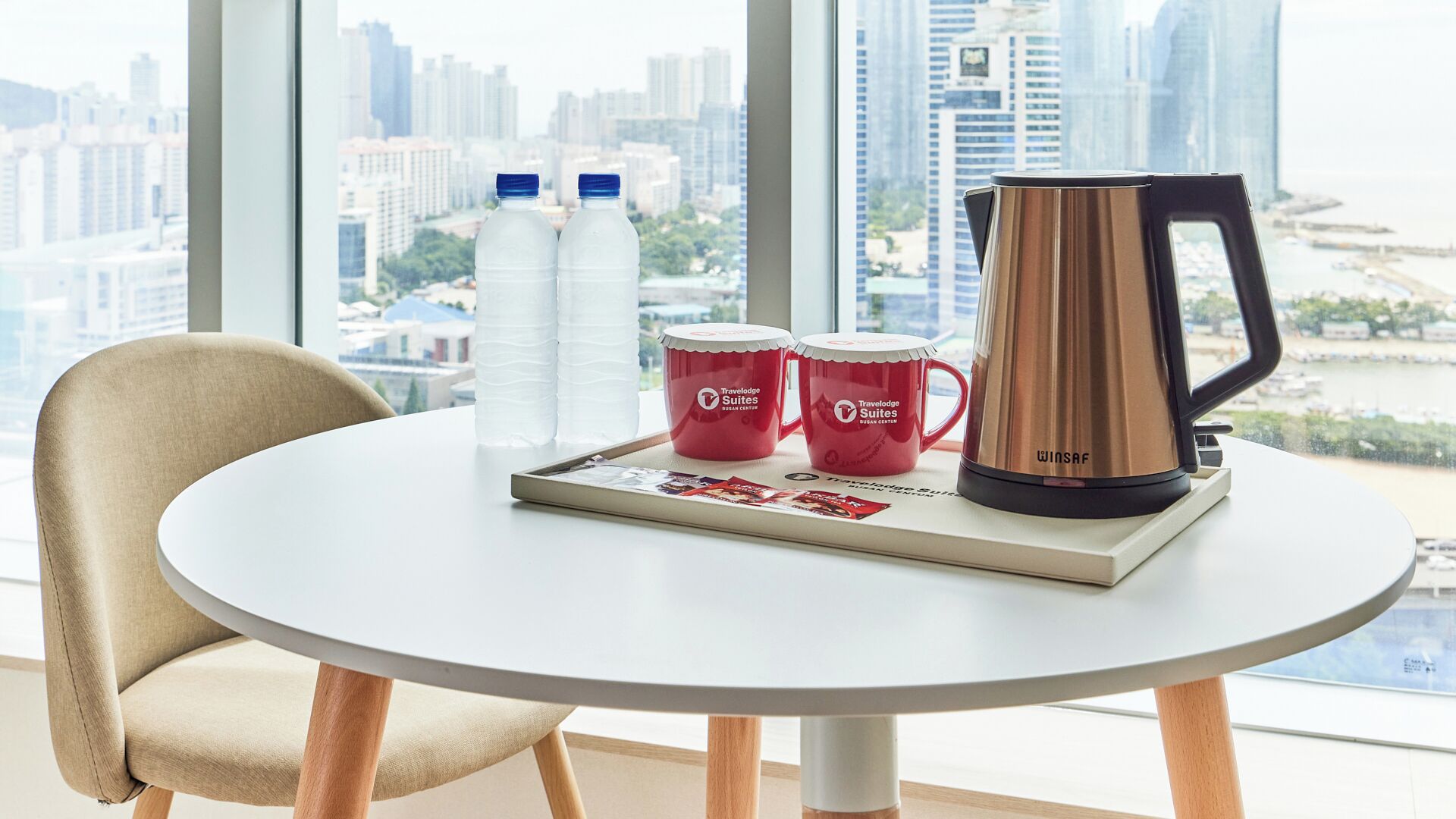 Uma mesa com uma cafeteira, duas canecas vermelhas e duas garrafas de água em um quarto com vista para a cidade.