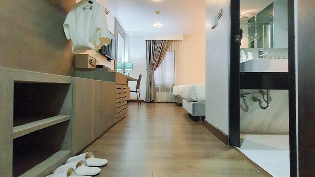 Um corredor de hotel com piso de madeira, uma área de estar com um sofá e uma poltrona, uma mesa pequena com luminária e um espelho na parede.