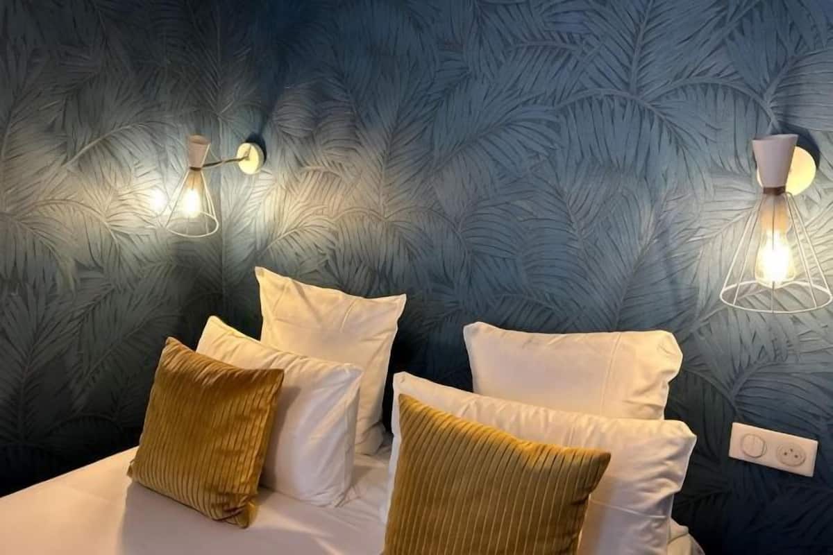 Quarto com papel de parede estampado, cama com almofadas brancas e douradas, e duas luminárias fixadas na parede.