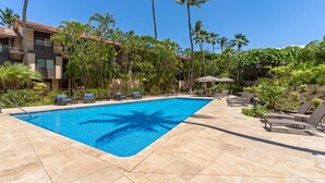 Piscina com espreguiçadeiras, cercada por palmeiras e um edifício com varandas.
