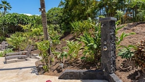 Uma área externa ajardinada com um conjunto de assentos curvos, um pilar de pedra e diversas plantas tropicais.