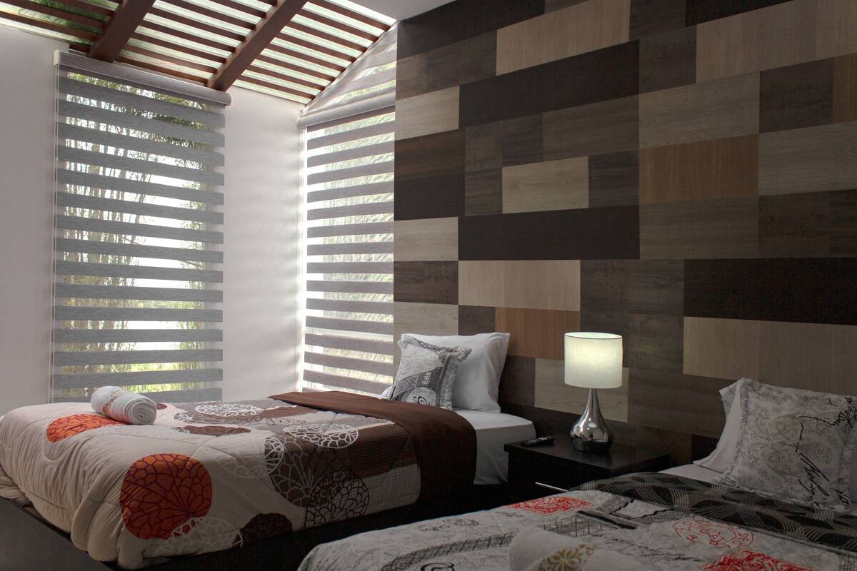 Quarto de hotel moderno com uma janela grande, uma cama com colcha estampada, uma mesa de cabeceira com abajur e uma parede com design de painel de madeira geométrica.