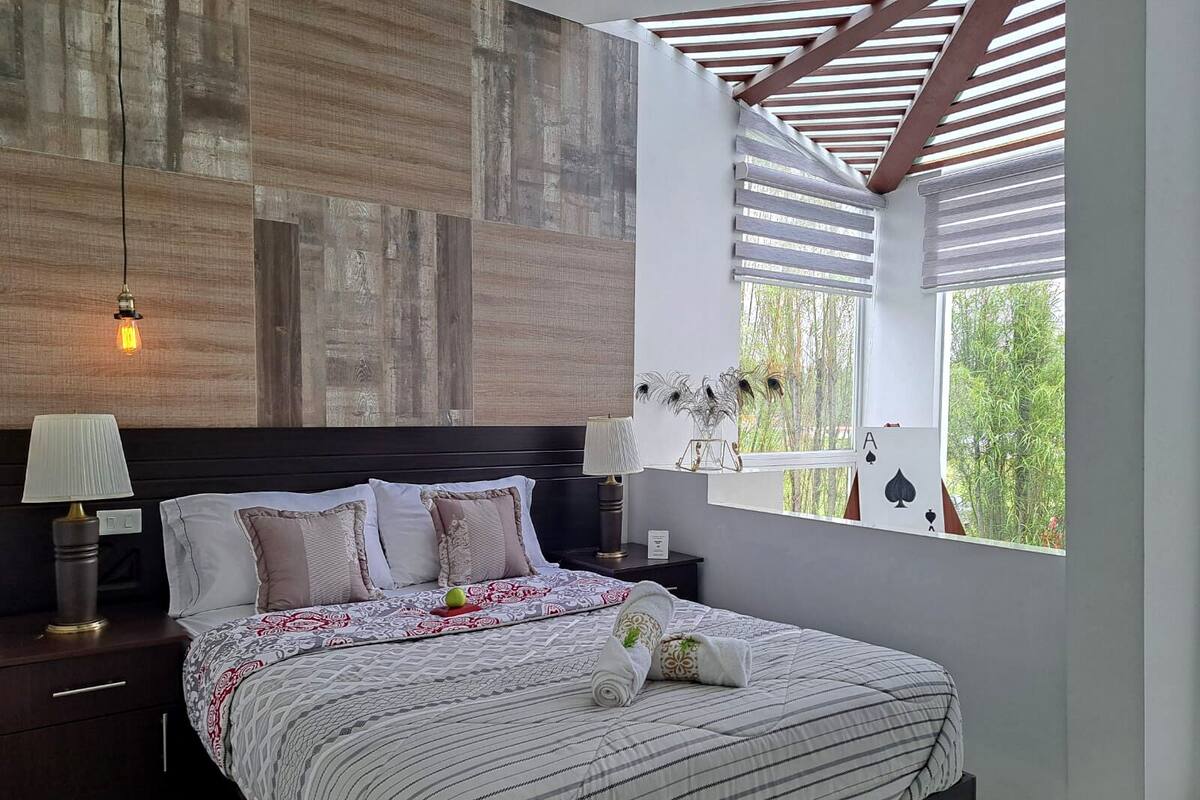 Um quarto moderno com uma cama grande, uma parede de destaque em madeira, uma janela com vista para o verde e um teto com vigas de madeira.