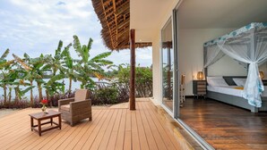 Quarto com cama de dossel, deck de madeira com telhado de palha e vista para palmeiras e o mar.