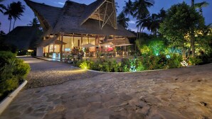 Um resort com um edifício de telhado de palha, um calçadão e vegetação tropical.