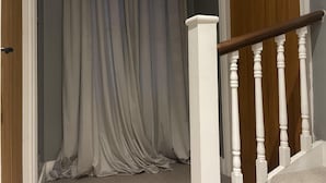 Uma escadaria com balaústres brancos e corrimão de madeira, conduzindo a um quarto com cortina cinza.