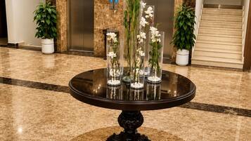 Um hall de entrada com piso de mármore, uma mesa redonda com tampo de vidro, plantas em vasos e dois elevadores.