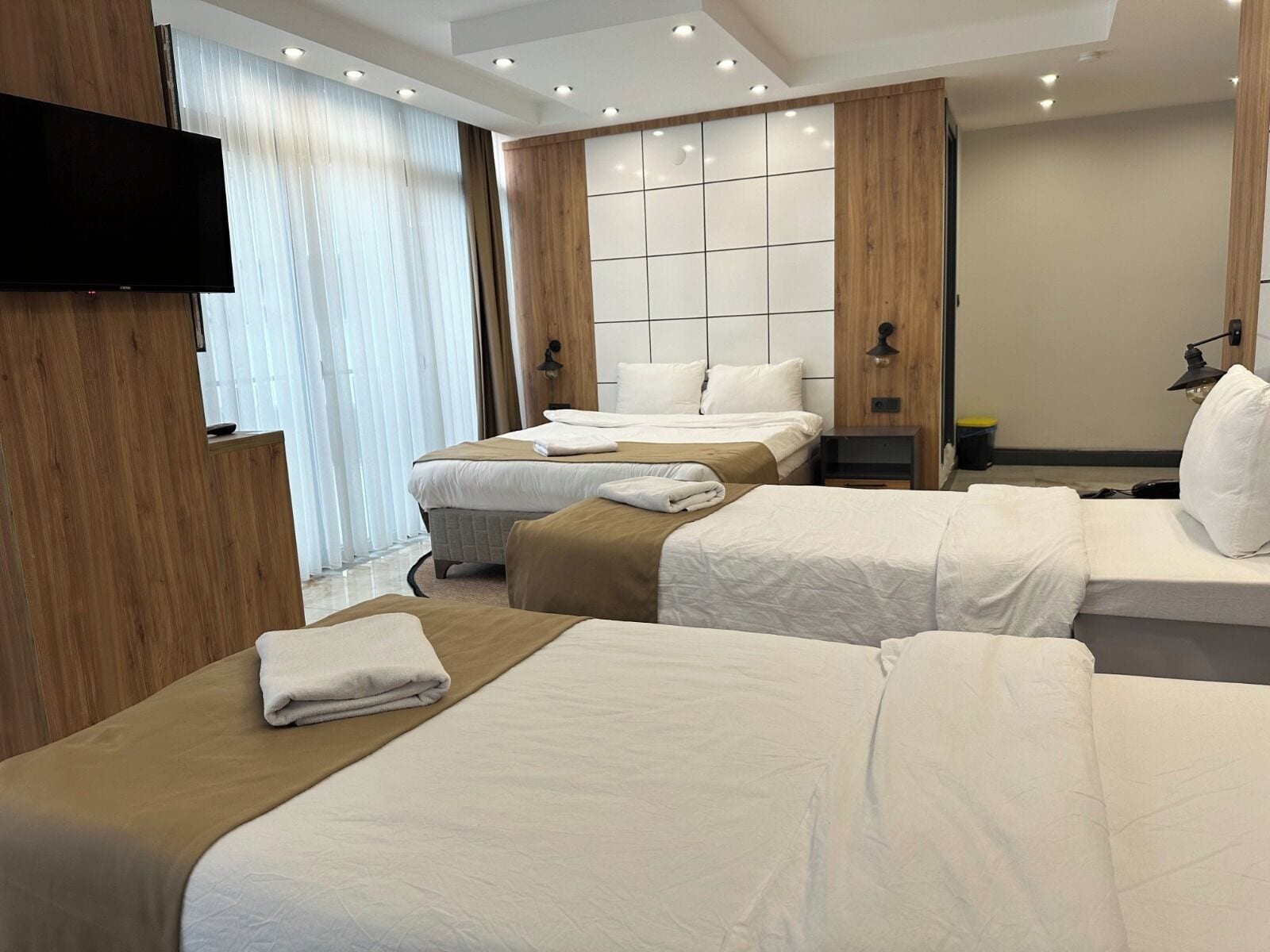 Quarto de hotel com três camas, televisão de tela plana na parede e uma grande janela com cortinas.