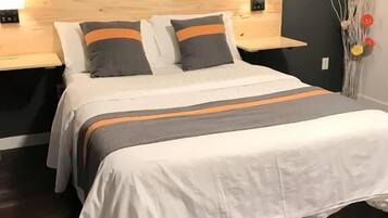 Quarto moderno com cabeceira de madeira, colcha branca com listras laranja e piso de madeira escura.