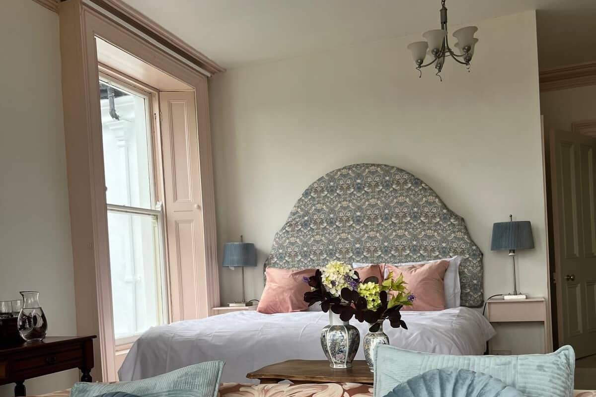 Quarto com cama grande, cabeceira decorativa, sofá estampado com flores e um vaso com flores.