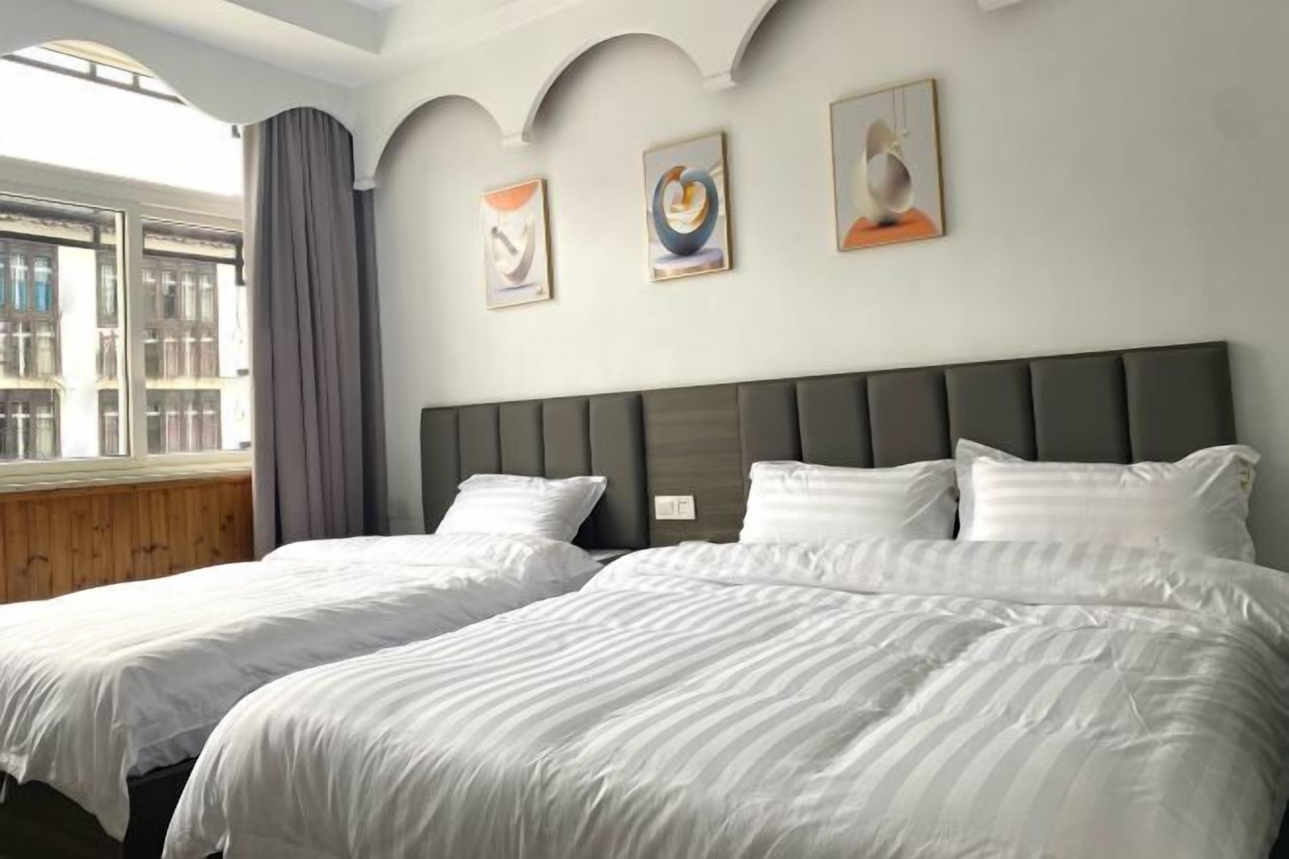 Quarto de hotel com duas camas, uma janela grande com cortinas e três quadros abstratos na parede.