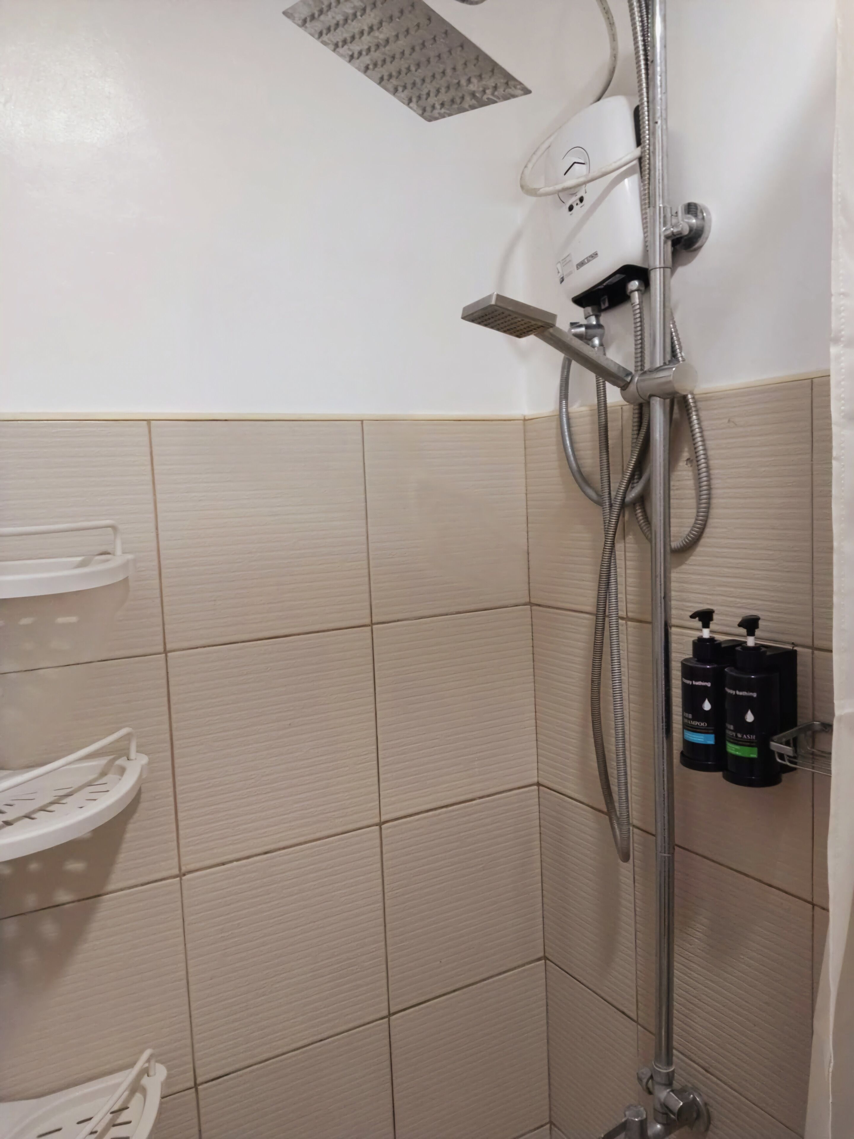 Área de chuveiro com chuveiro manual, painel de controle fixado na parede e dois frascos de shampoo em uma prateleira.
