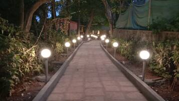 Um caminho iluminado por luzes uniformemente espaçadas, ladeado por vegetação e árvores, conduz a um edifício ao longe.