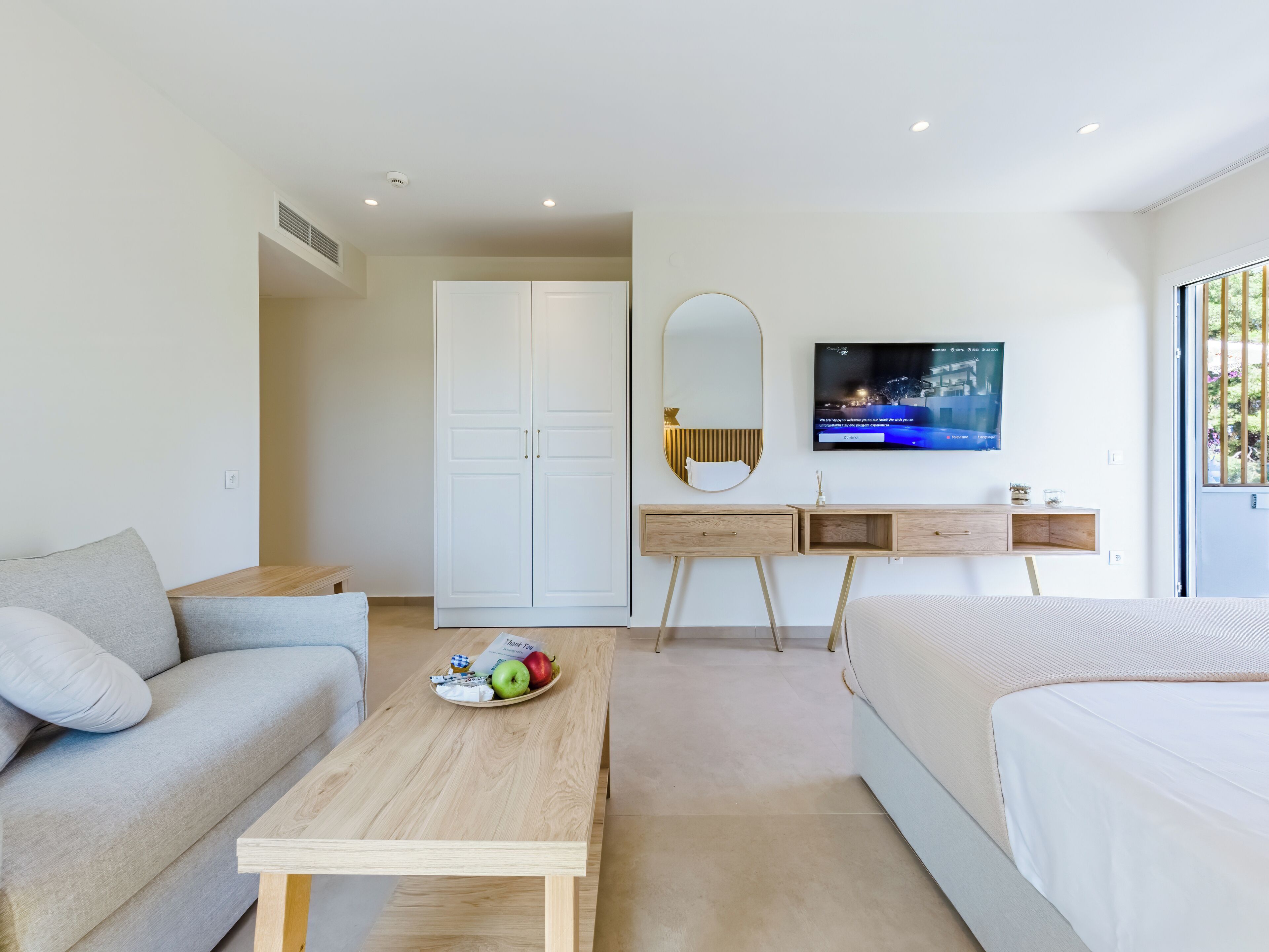 Quarto de hotel moderno com sofá, mesa de jantar com frutas, televisão, escrivaninha e guarda-roupa.