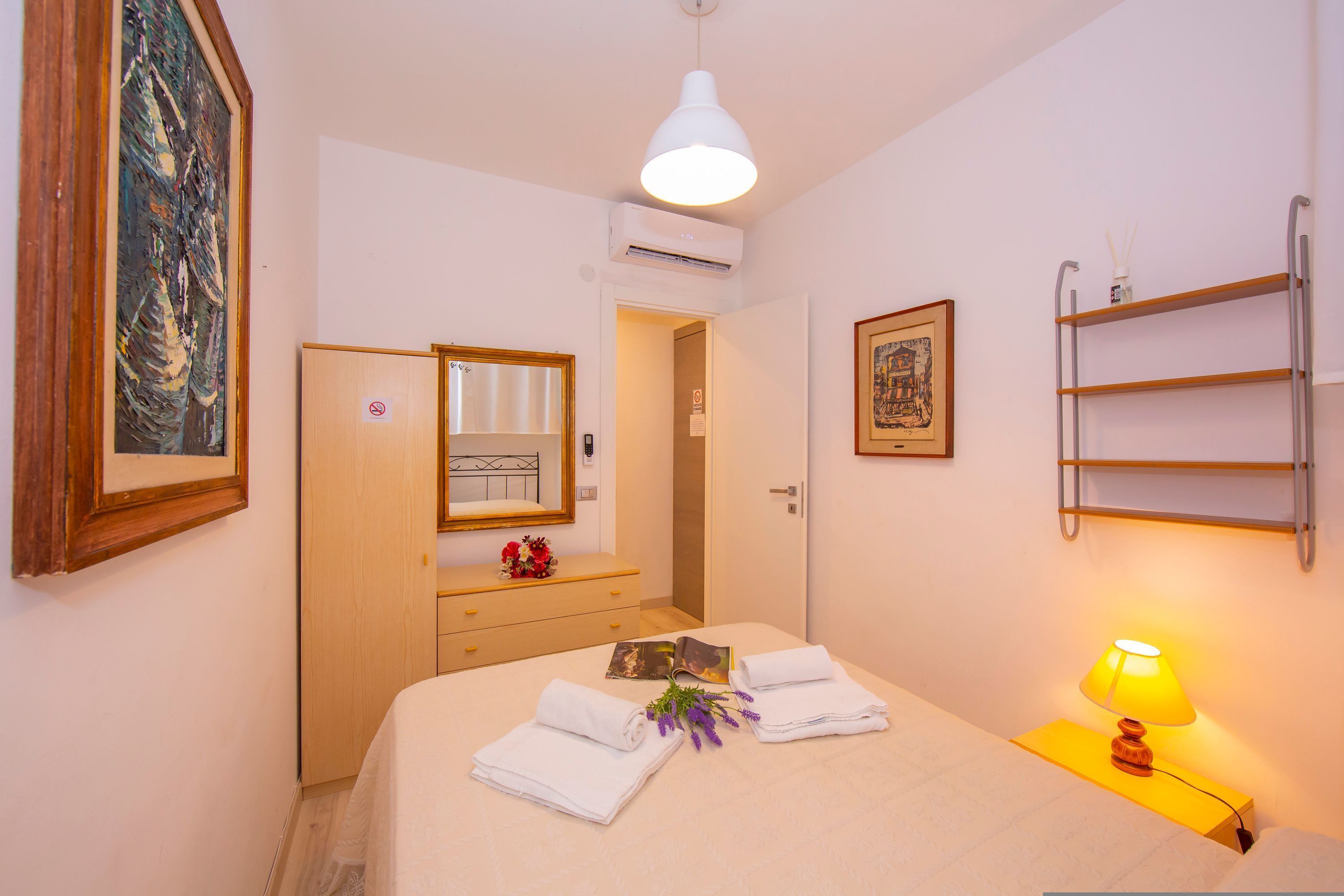 Cama bem arrumada com lençóis brancos, abajur, quadro na parede e cômoda de madeira com espelho.