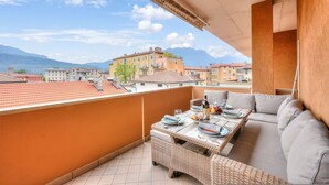 Varanda com mesa posta para refeições, mobiliário de vime e vista para edifícios e montanhas.