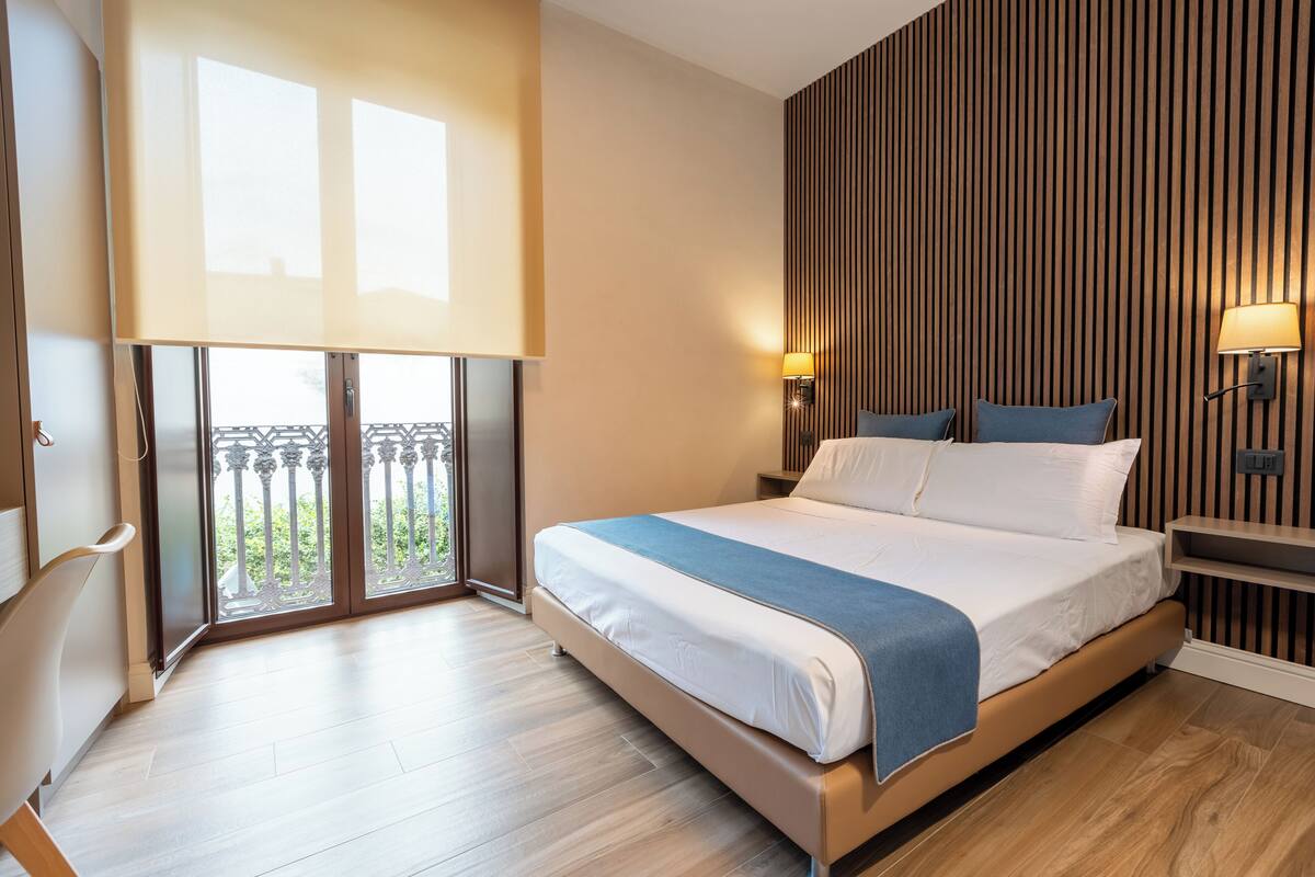 Quarto de hotel moderno com uma cama grande, uma varanda com guarda-corpo e duas luminárias fixadas na parede.