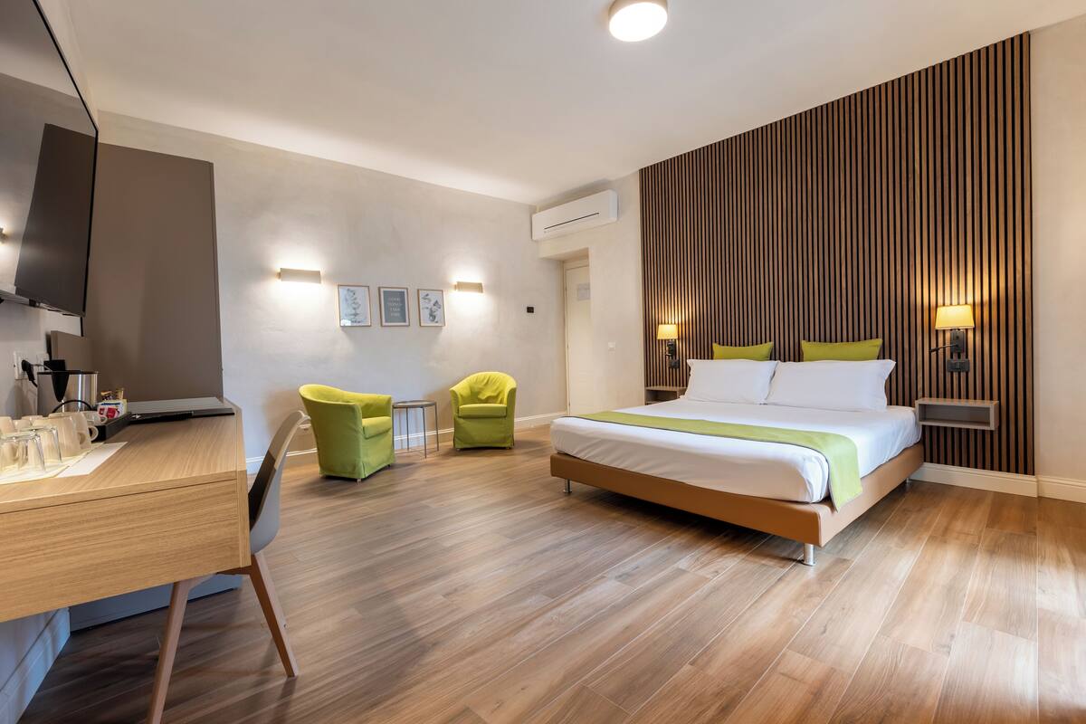 Quarto de hotel moderno com piso de madeira, cama com roupa de cama verde, escrivaninha com laptop, poltrona verde e TV fixada na parede.