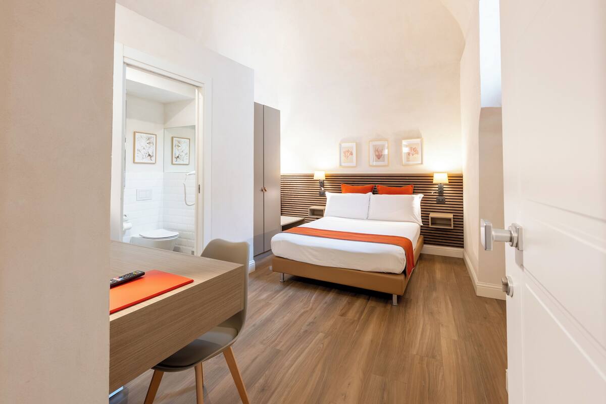 Quarto de hotel com cama, mesa de cabeceira, cadeira e banheiro visível por uma porta aberta.