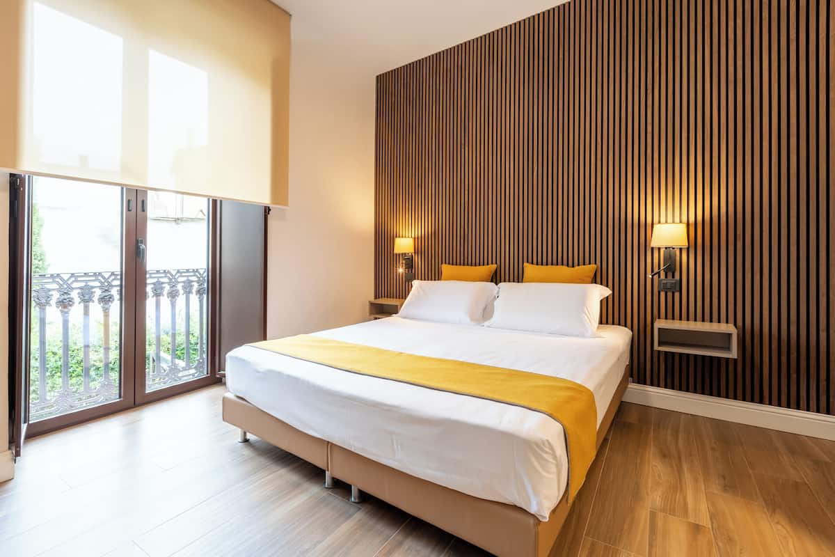 Quarto de hotel moderno com uma cama grande, painel de parede de madeira, duas luminárias de cabeceira e uma varanda com guarda-corrimão.