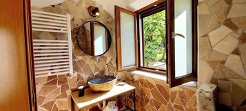 Um banheiro com parede de pedra, um espelho redondo, pia com uma tigela decorativa e janela com vista para o verde.