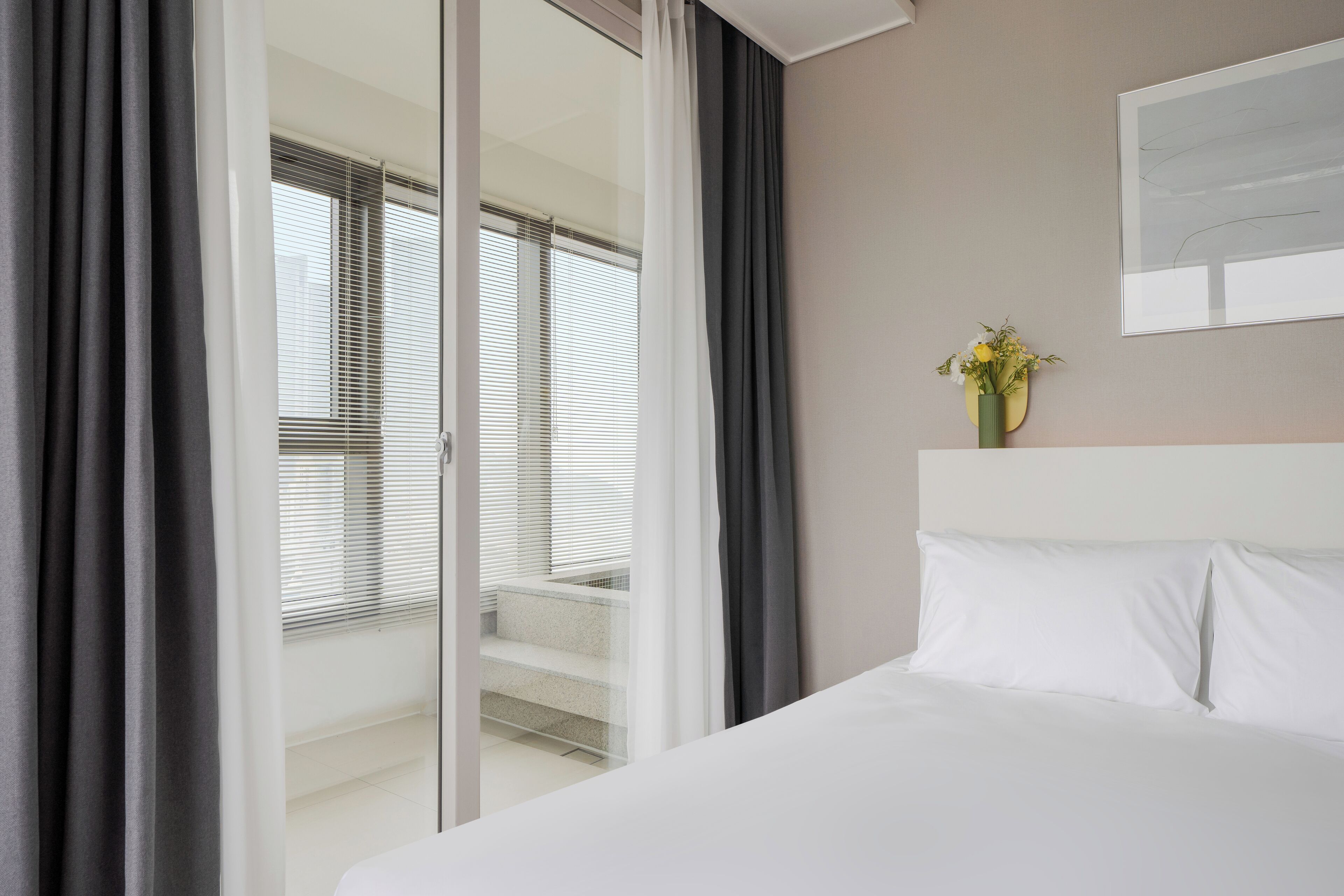 Quarto de hotel com uma cama grande, um vaso com flores e vista para uma varanda com persianas.