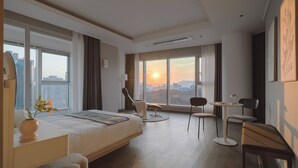 Quarto de hotel moderno com uma cama grande, uma mesa, uma cadeira, uma mesinha de apoio e vista para a cidade ao pôr do sol.