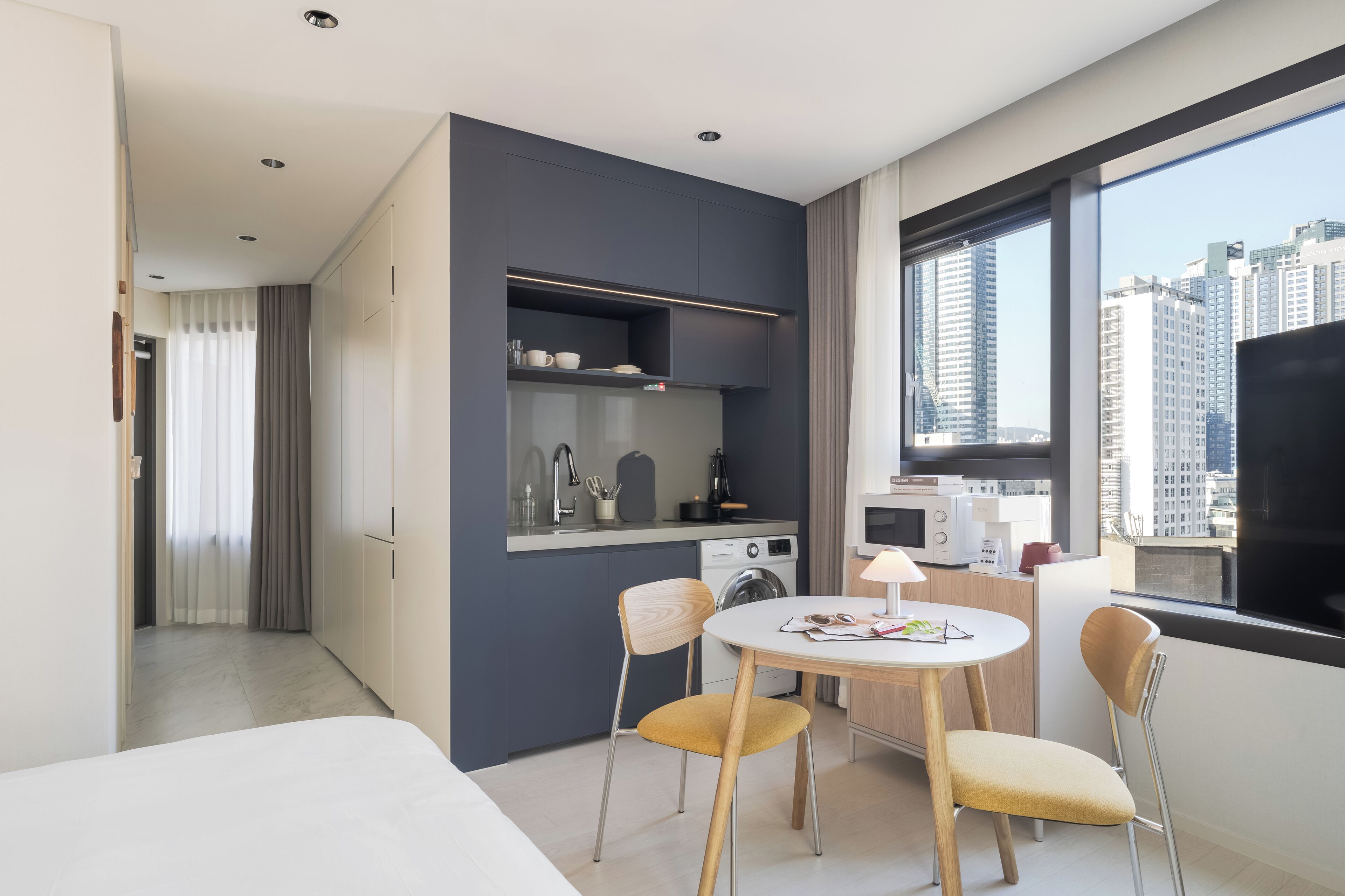 Quarto de hotel moderno com área de refeições, cozinha compacta e uma grande janela com vista para a cidade.