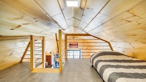 Quarto de sótão com pé-direito alto, escada de ripas de madeira, cama com edredom listrado e claraboia.