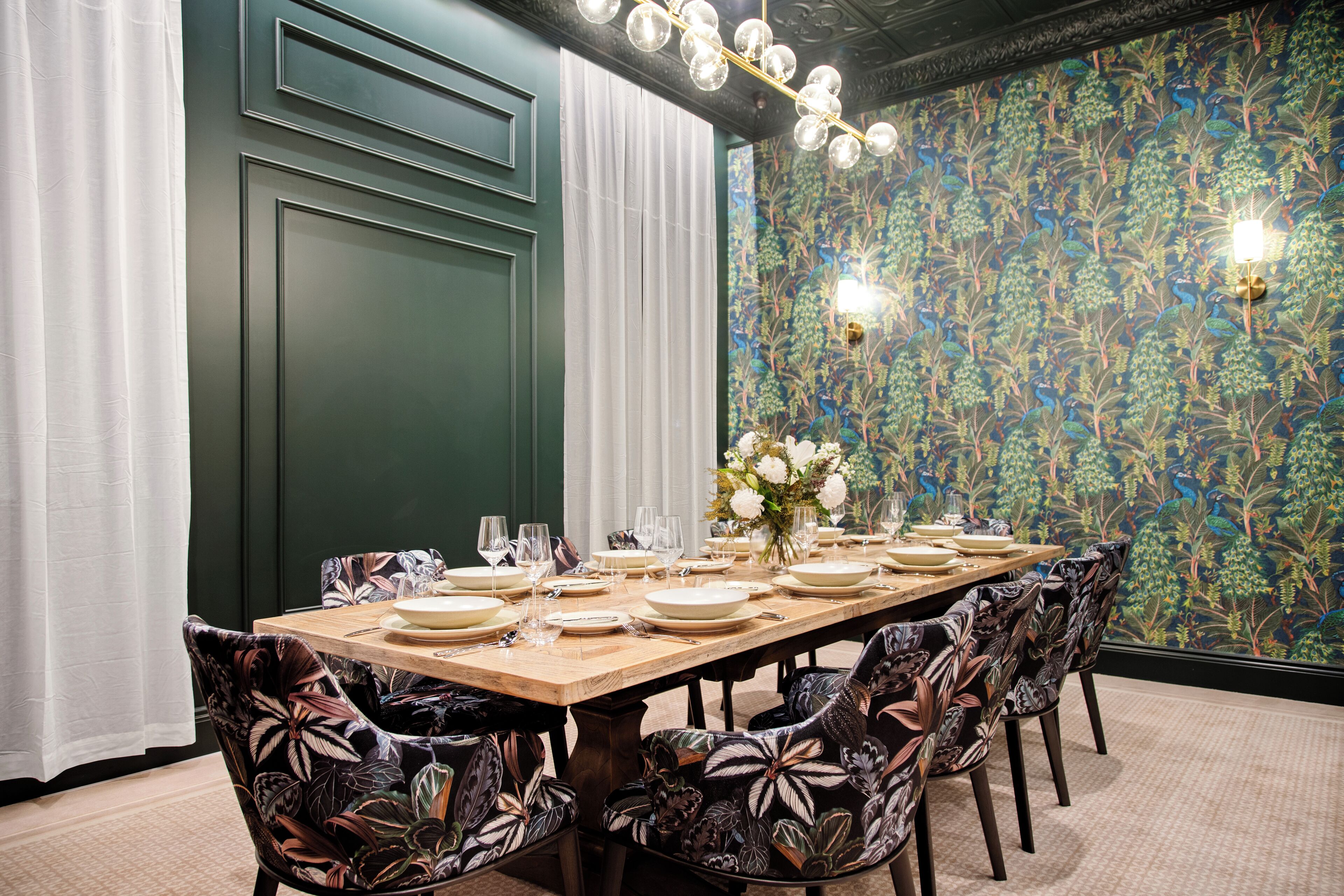 Uma sala de jantar com uma longa mesa de madeira posta para seis pessoas, rodeada por cadeiras com padrão floral.