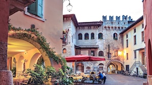 Um café ao ar livre com mesas e cadeiras, rodeado por edifícios de pedra e uma estrutura semelhante a um castelo ao fundo.