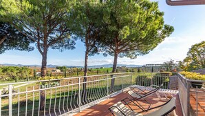 Terraço com guarda-corrimão de metal, chão de azulejos e vista para uma paisagem com árvores e colinas ao longe.
