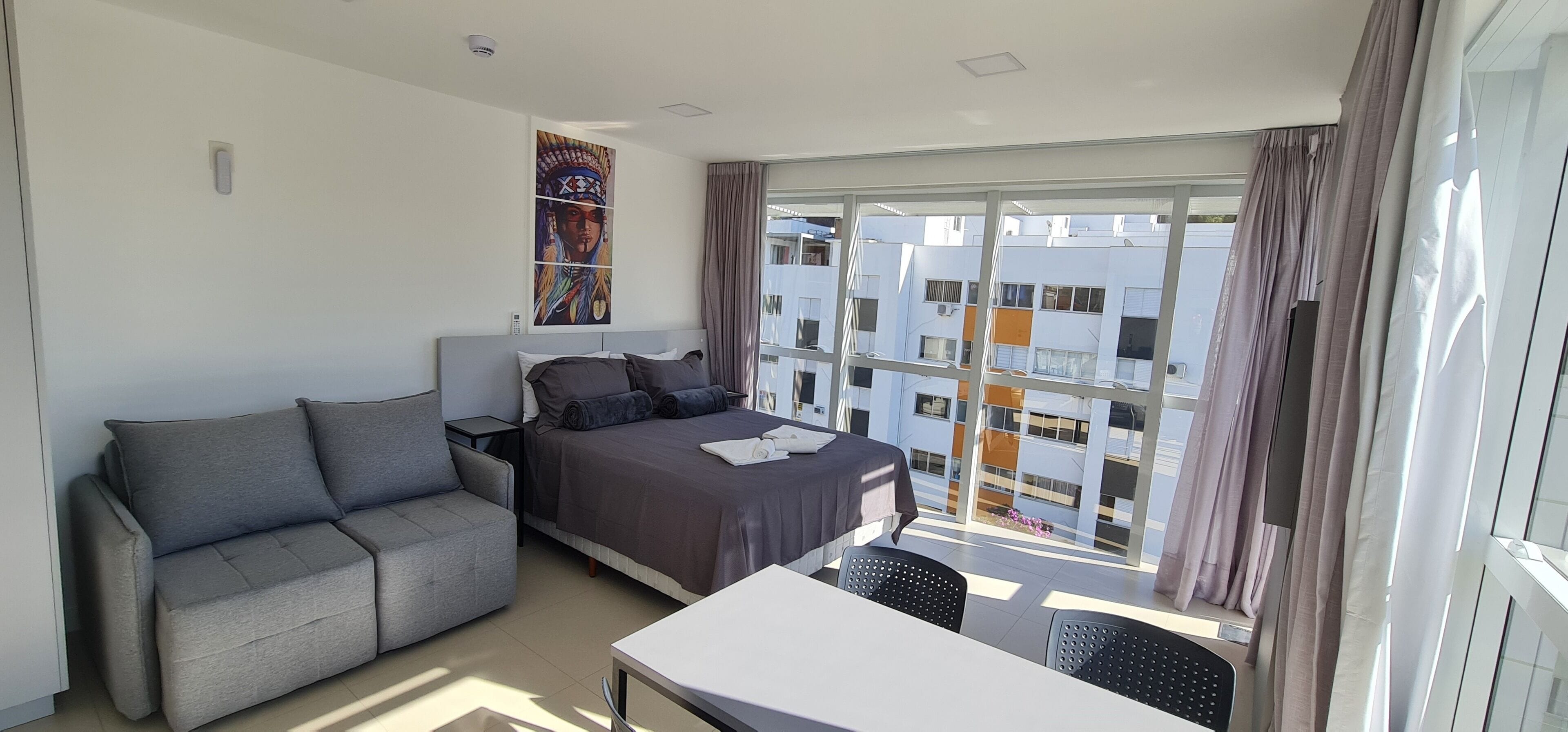 Quarto de hotel moderno com cama, sofá cinzento e área de refeições.